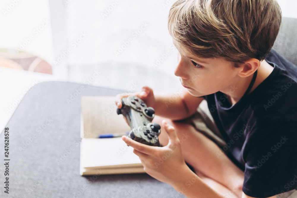 Blond child playing video games instead of studying sitting home sofa ...