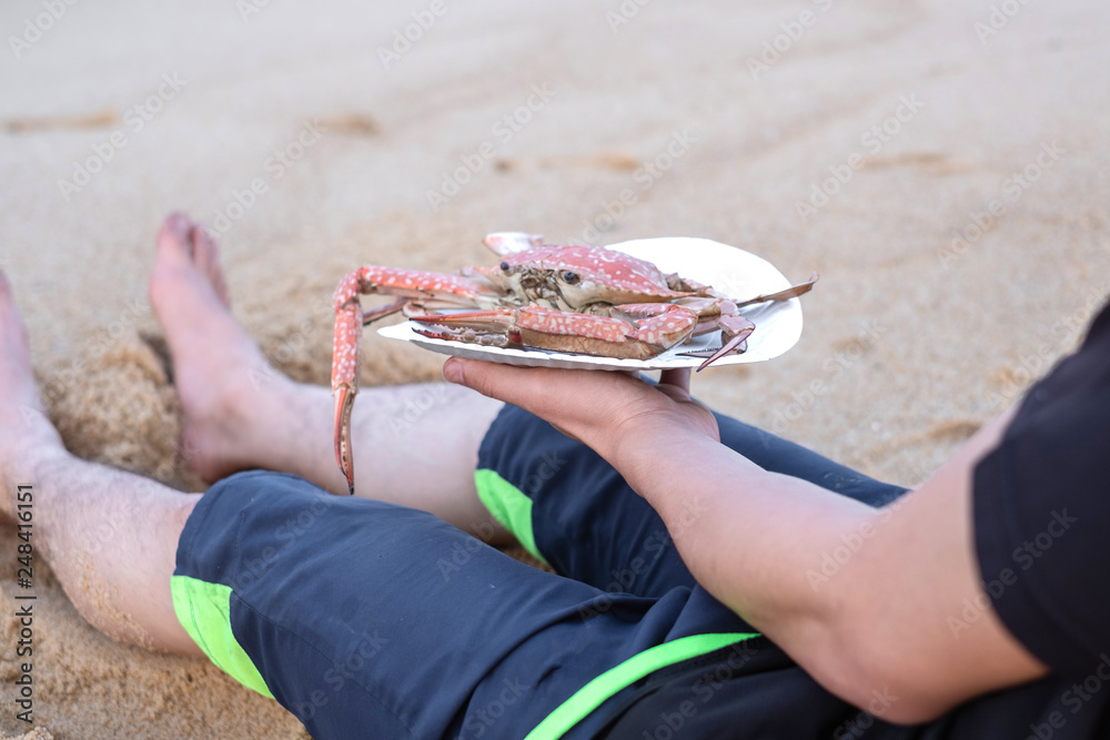 Young man holding cooked crab in a whote paper plate sitting on beach ...