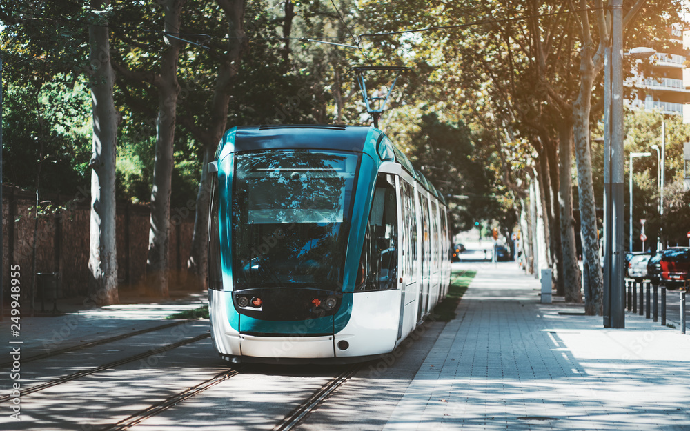 A modern neat tram in city alleyway surrounded by trees; blue and white ...