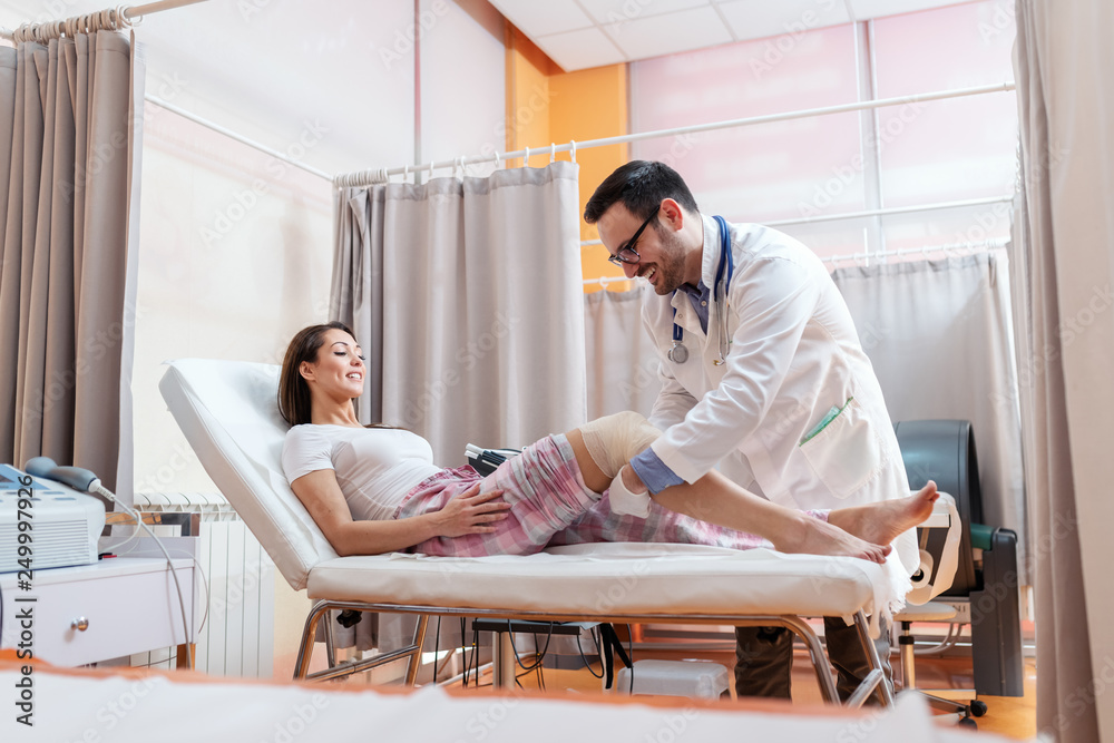 Smiling polite doctor folding knee while his patient laying down on ...