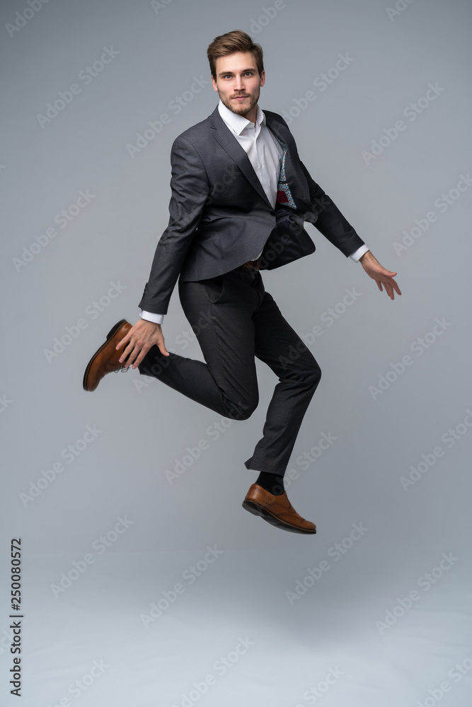 Mid-air style. Handsome young man in full suit jumping against gray ...