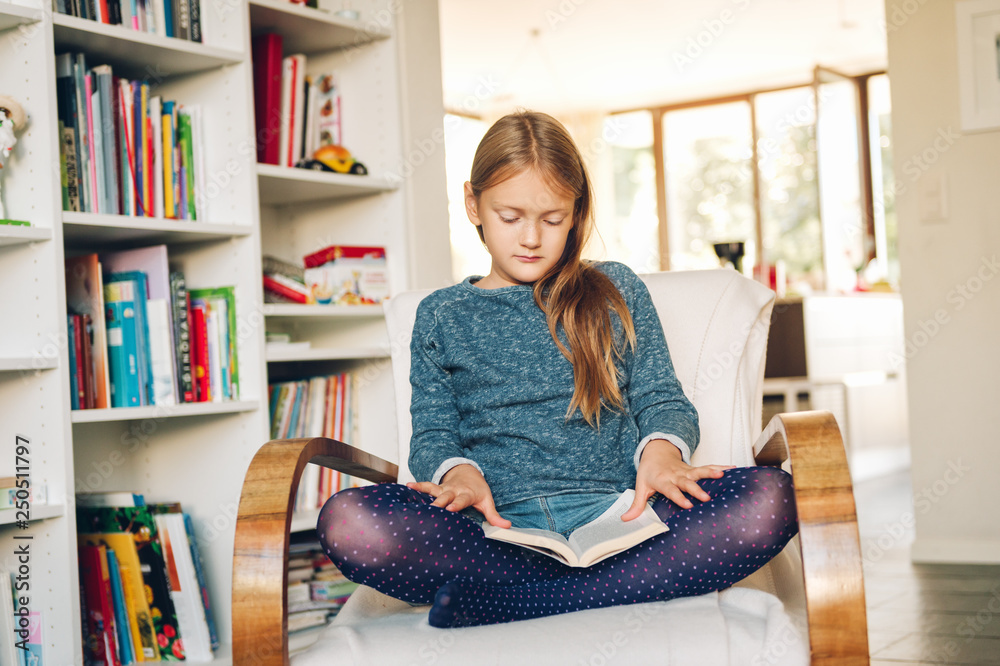 Cute little girl sitting in a white chair at home and reading a book ...