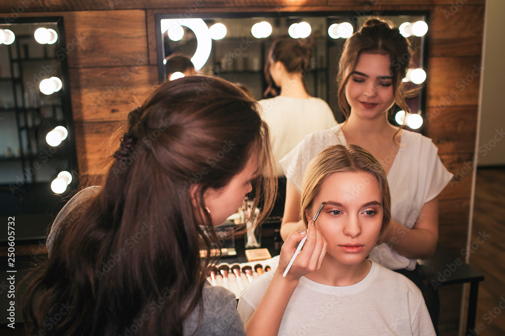 Serious blonde woman sit on chair in beauty room. Make up artist put ...