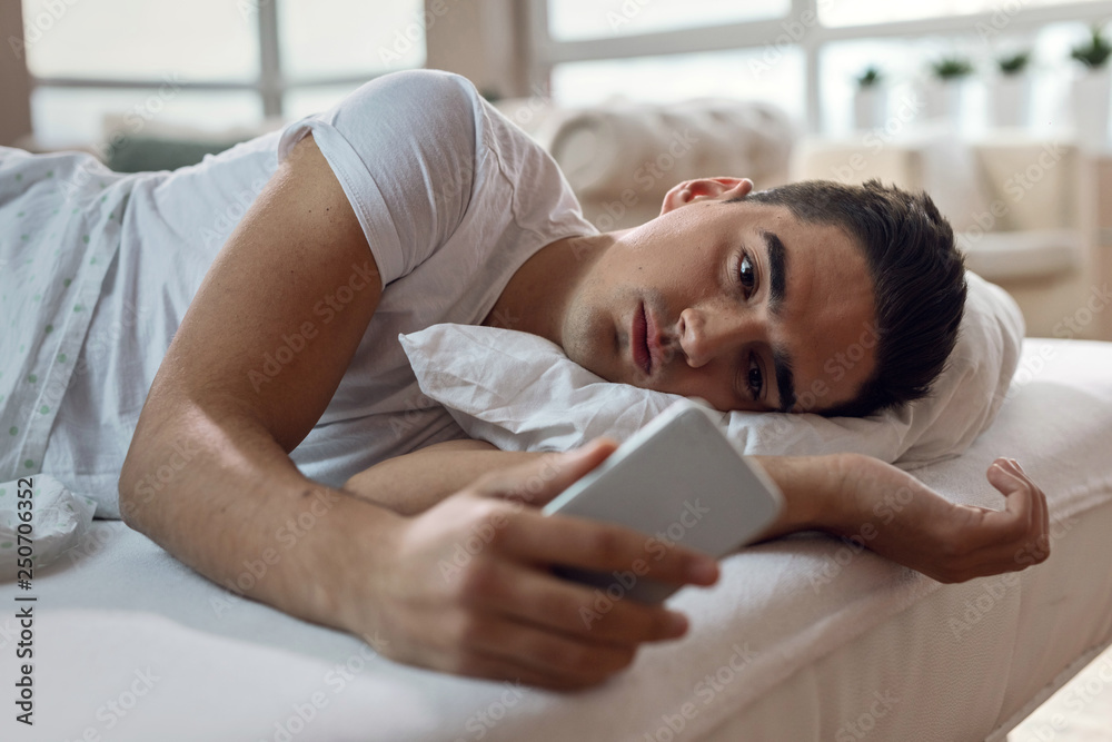 Young Sad Man Lying On Bed And Reading Text Message On Mobile Phone