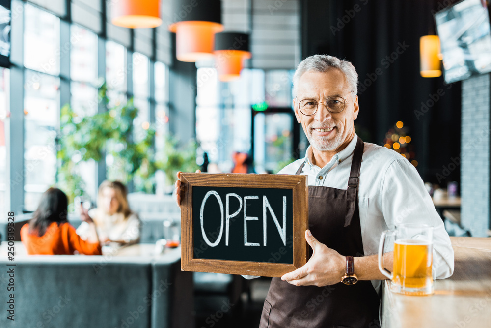 smiling owner of pub holding open sign and standing near bar counter ...