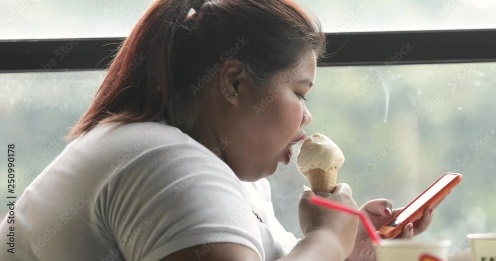 Overweight woman eating ice cream cone while using a mobile phone in ...