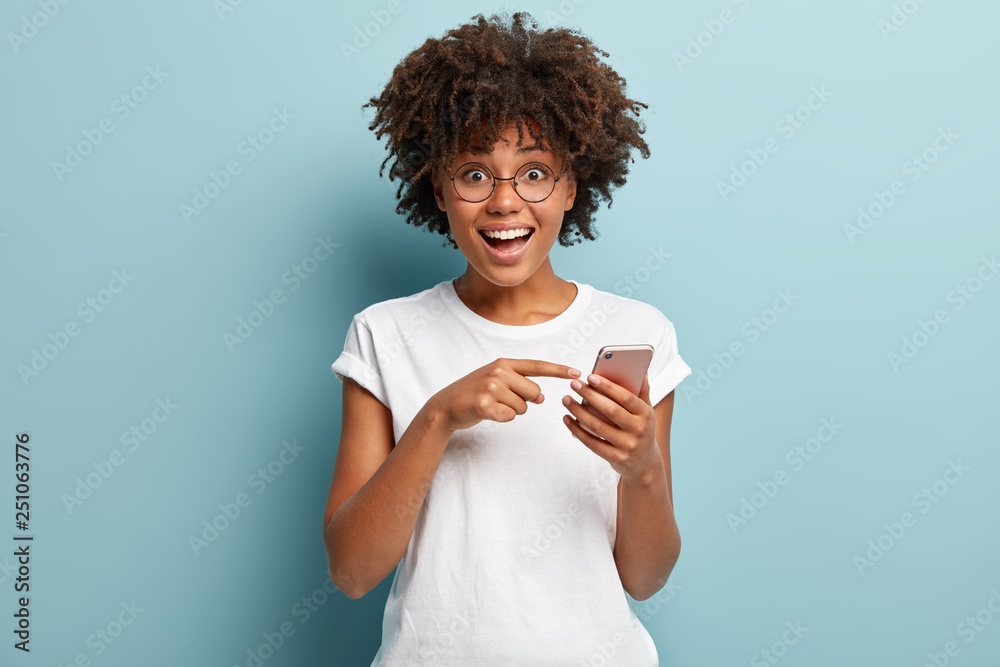Joyous African American lady with big smile, points at screen of cell ...
