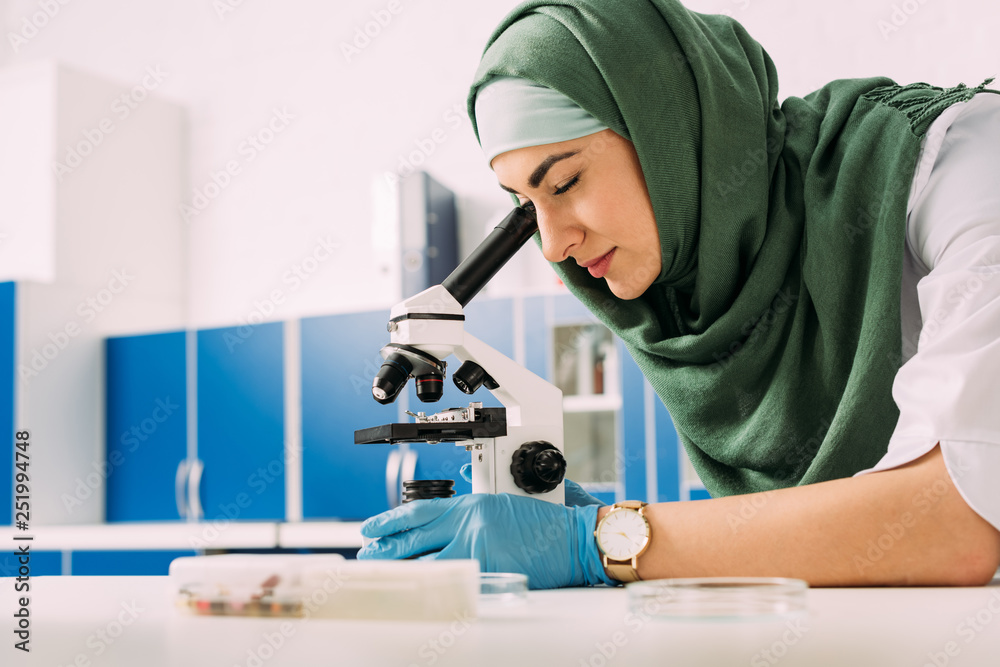 concentrated female muslim scientist looking through microscope during ...