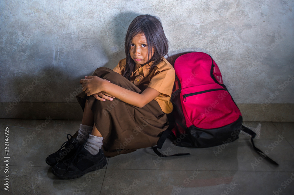 sad and depressed 8 or 9 years old child in school uniform sitting on the floor with her bad ...