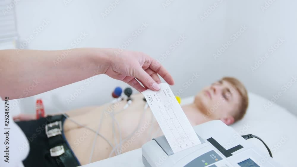 A medical clinic. A young man receiving a ECG procedure. A results tape ...
