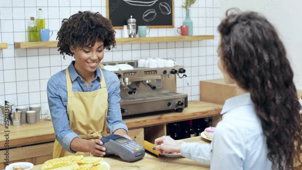 beautiful african american female cashier holding terminal while female ...