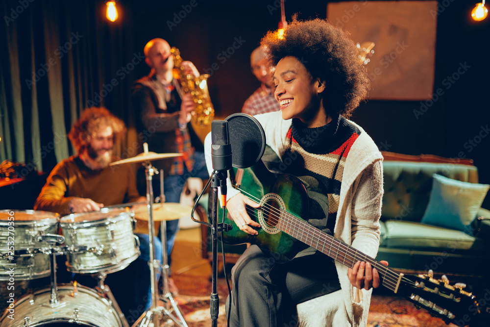 Mixed race woman singing and playing guitar while sitting on chair with ...