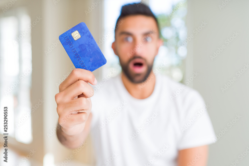 Handsome hispanic man holding credit card scared in shock with a ...