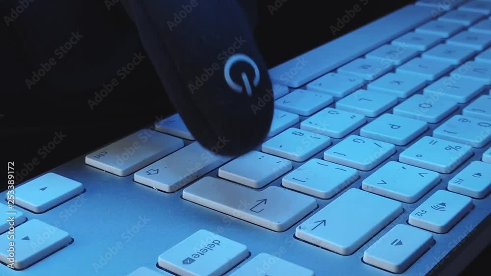 An eery blue light illuminates a keyboard in close up as a gloved ...