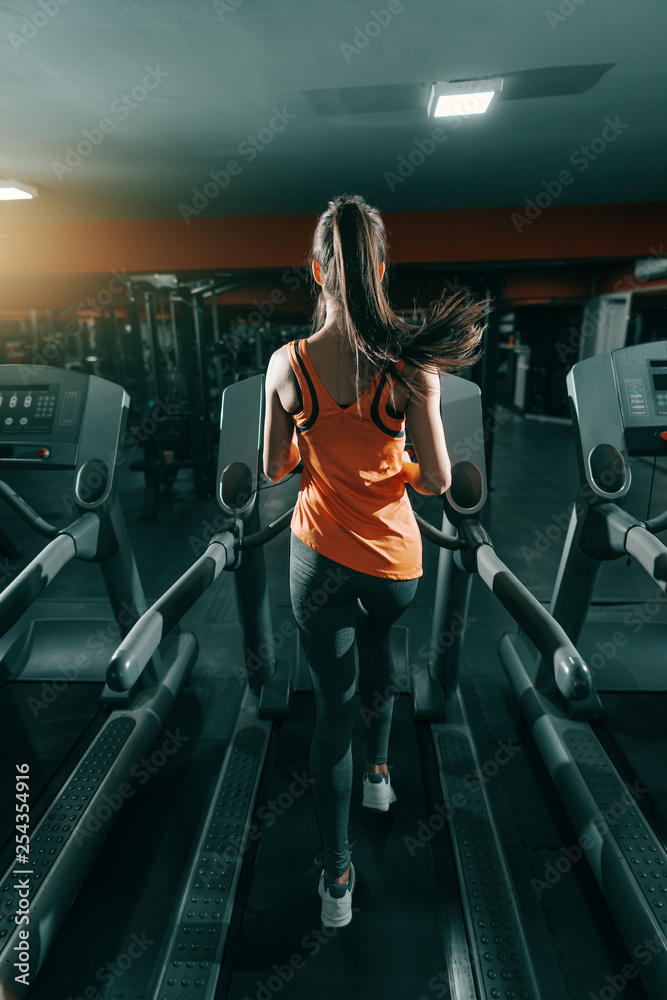 Slim brunette in orange sportswear and ponytail running on treadmill in ...