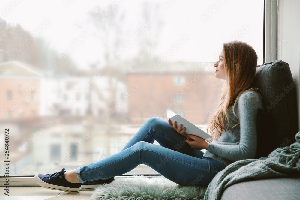 Beautiful young woman reading book while sitting on window sill Stock ...