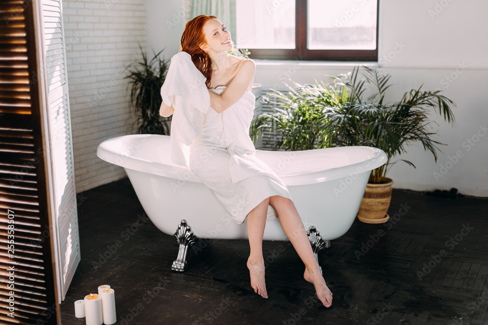 Young happy woman with ginger long hair is sitting on the edge of a bathtub and wiping her hair ...