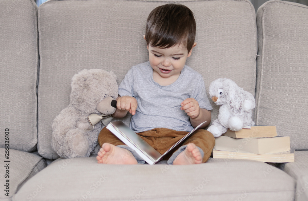 Cute smiling little baby boy reading book sitting on sofa with teddy ...