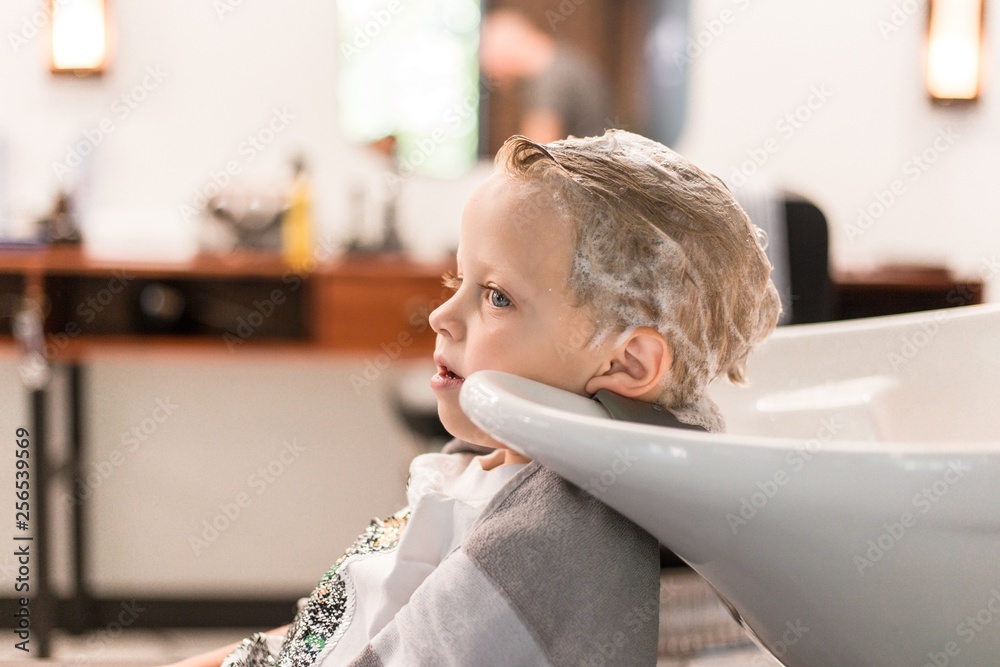 Little blond boy sitting in a Barber chair with soapy hair tipping his ...
