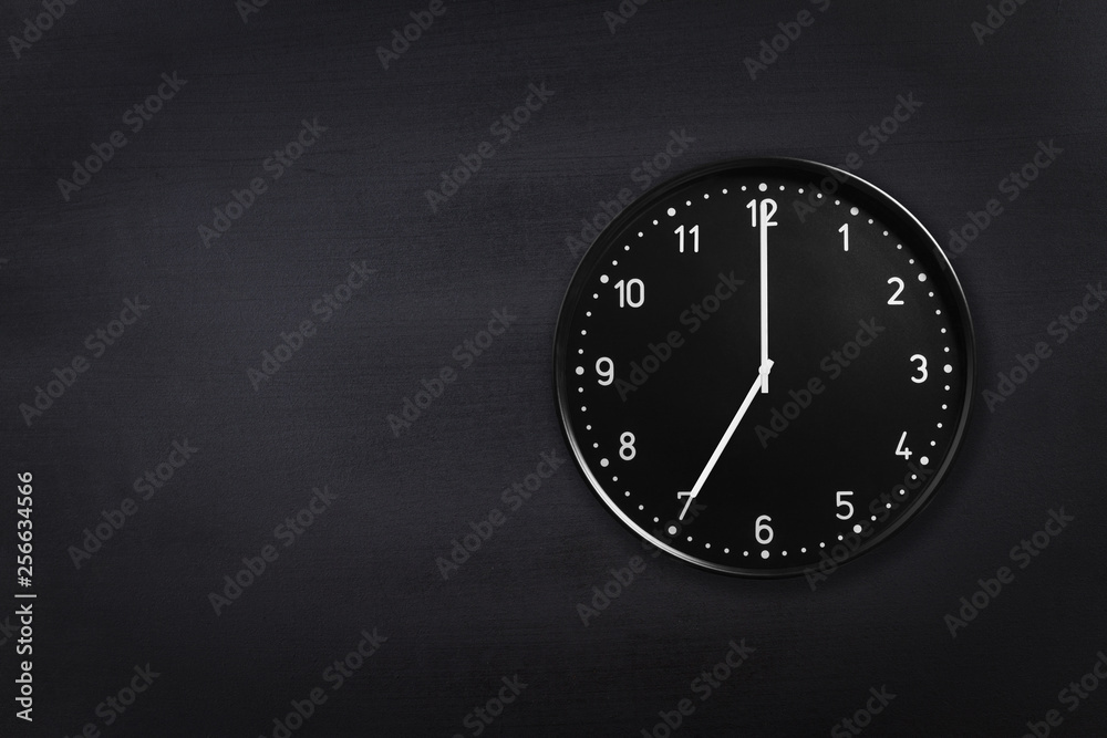 Black wall clock showing seven o'clock on black chalkboard background ...