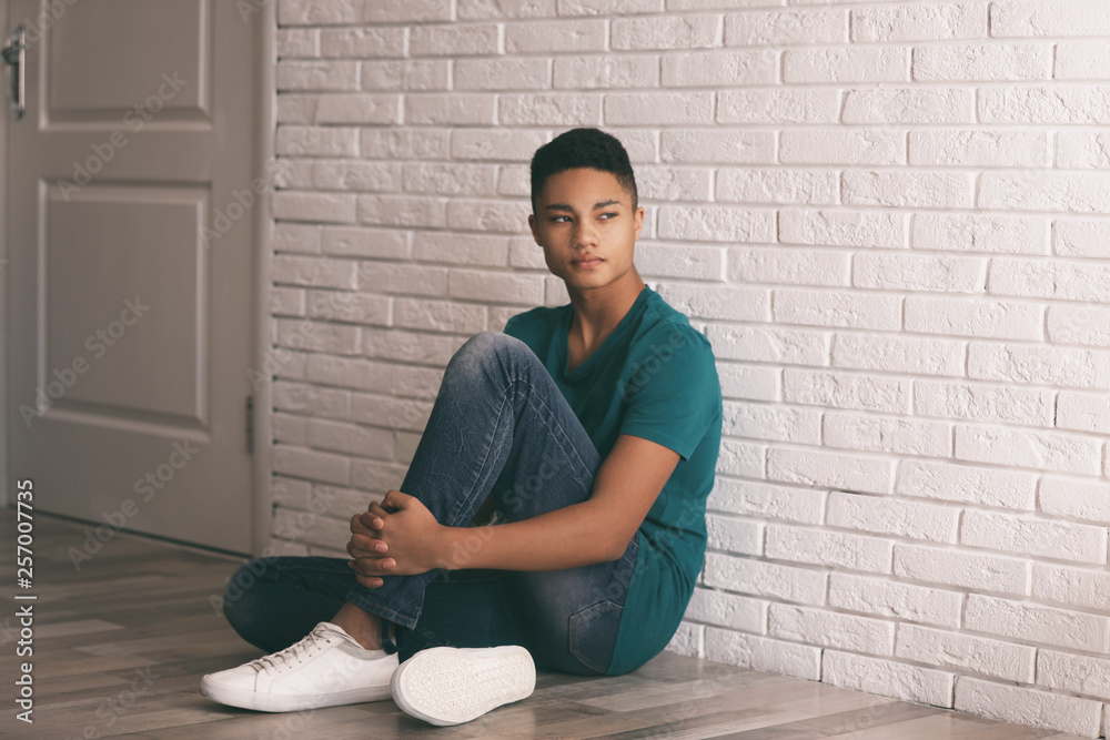 Upset African-American teenage boy sitting alone on floor near wall ...