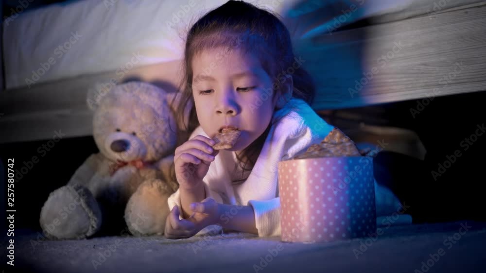 Happy child girl secretly eating candy under the bed in a dark bedroom ...