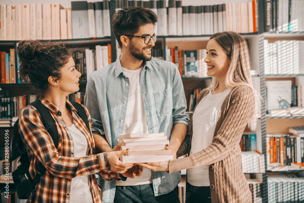 Three cheerful students standing in library and holding books they need ...