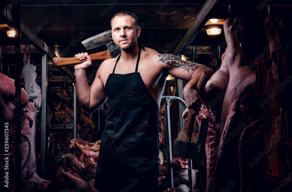 Shirtless tattooed butcher in apron posing with ax in a refrigerated ...