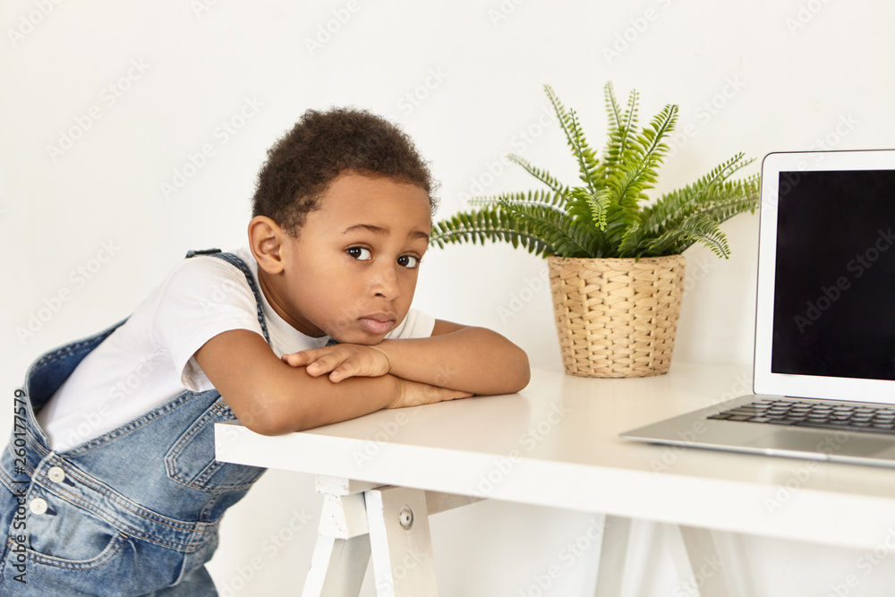 Sad upset Afro American preschooler placing elbows on white table with ...