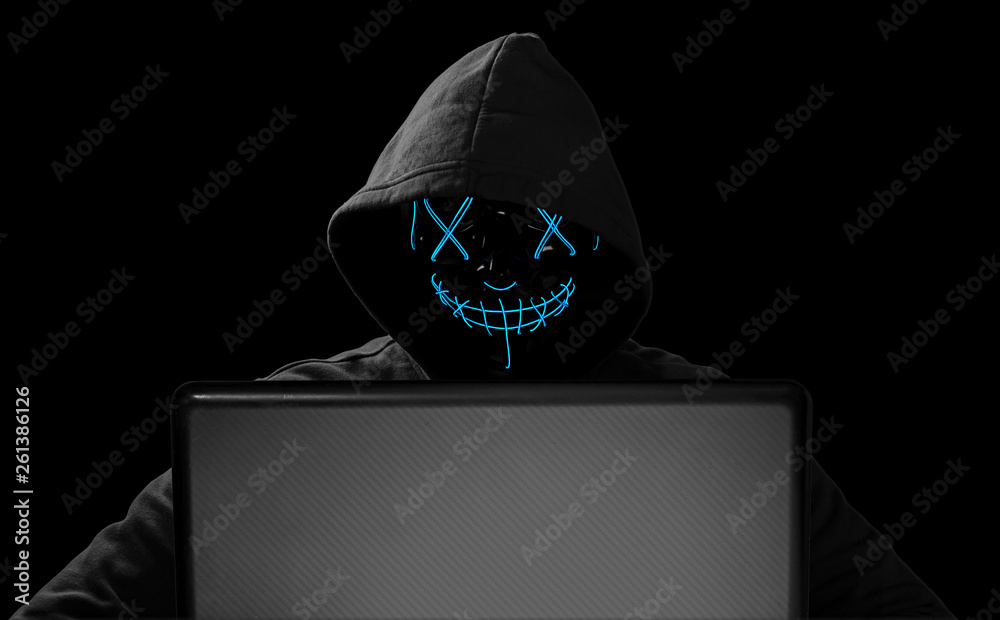 hacker with glowing mask behind notebook laptop in front of isolated black background internet ...