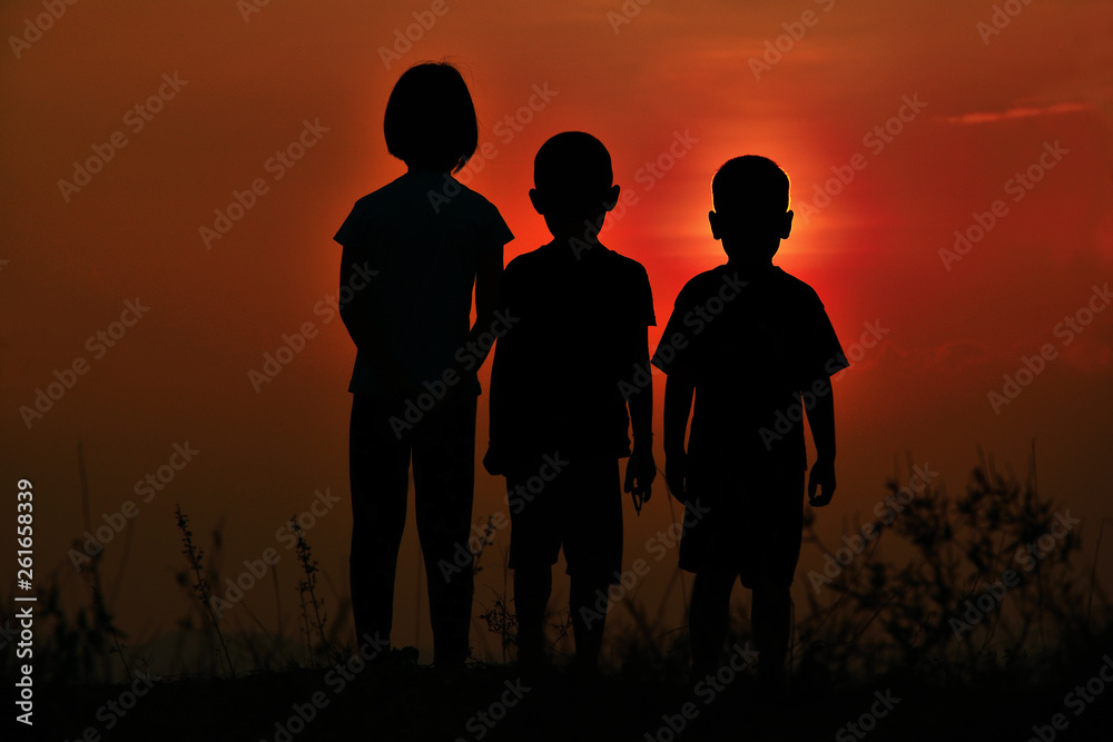 Black silhouette of three children standing together. There is a sky at ...