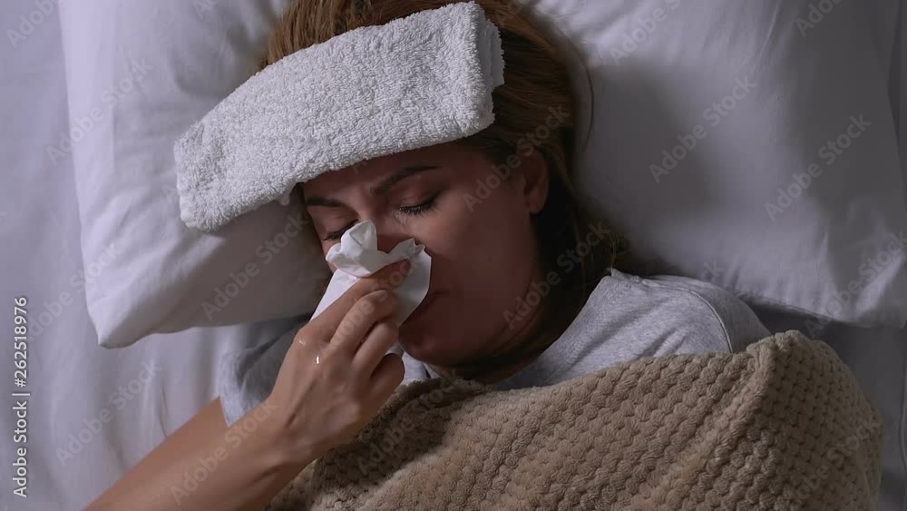 Sick woman with towel on forehead lying in bed, coughing and blowing ...
