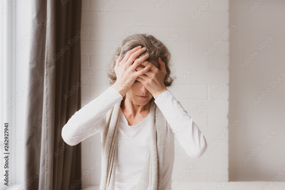 Middle aged woman with grey hair and scarf holding head with both hands ...