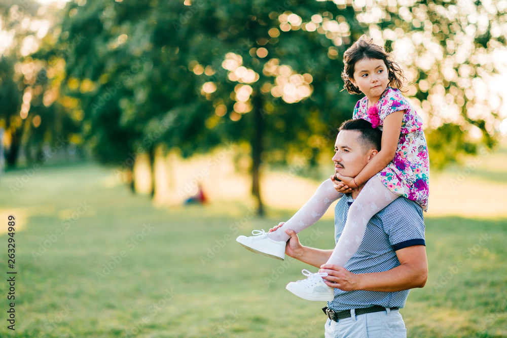 Dad carrying his daughter on shoulders in summer park at sunset. Father have fun with his little ...