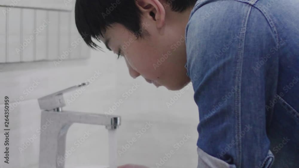 Preteen Asian boy washing his face by water from new faucet in bathroom. Young boy cleaning face ...