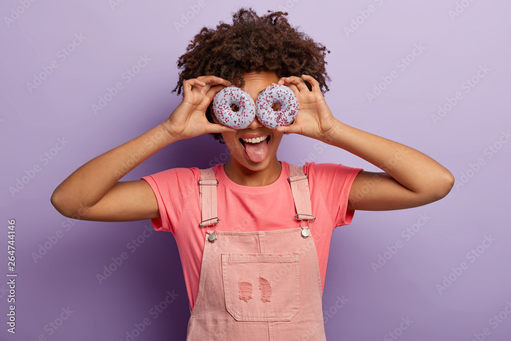 Positive female holds two purple doughnuts on eyes, sticks out tongue ...