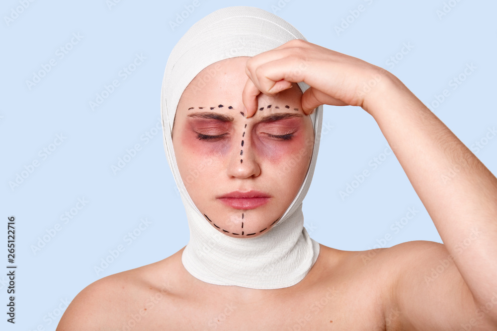 A beautiful young woman stands against blue wall with bandage on her ...
