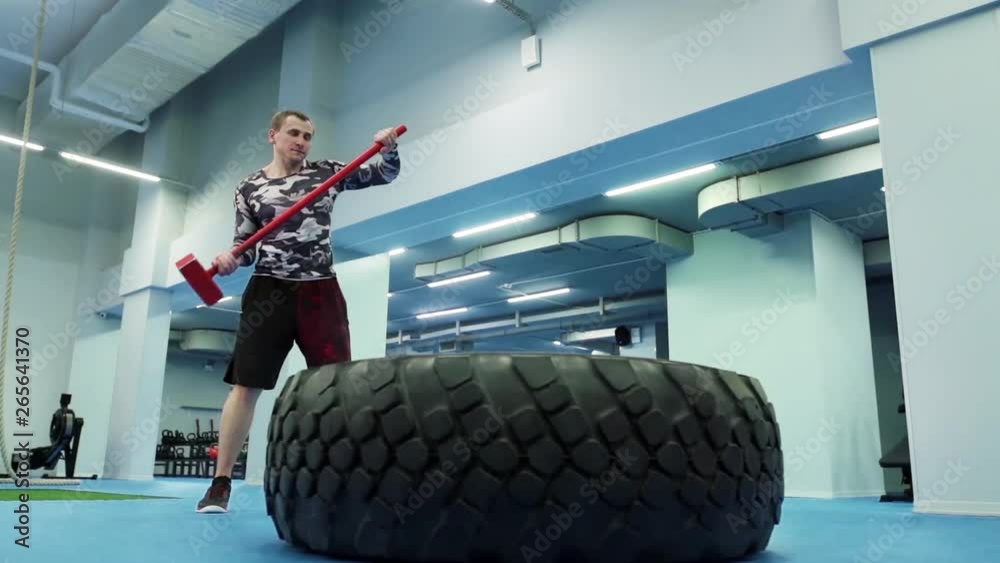 Muscular strong man with a hammer hits the huge wheel in the gym in ...