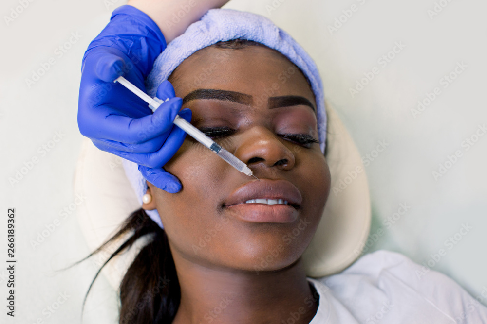 Beautiful young African woman gets cosmetic injection on her lips, isolated over light ...