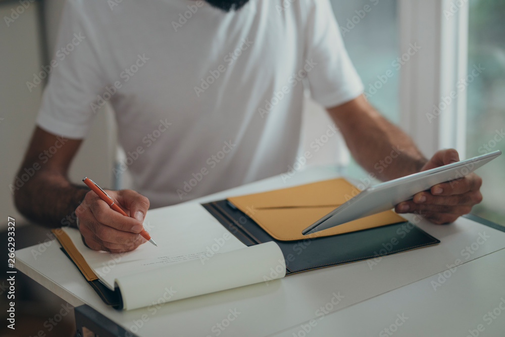Hands of unrecognisable man holding tablet and writing in his notebook ...