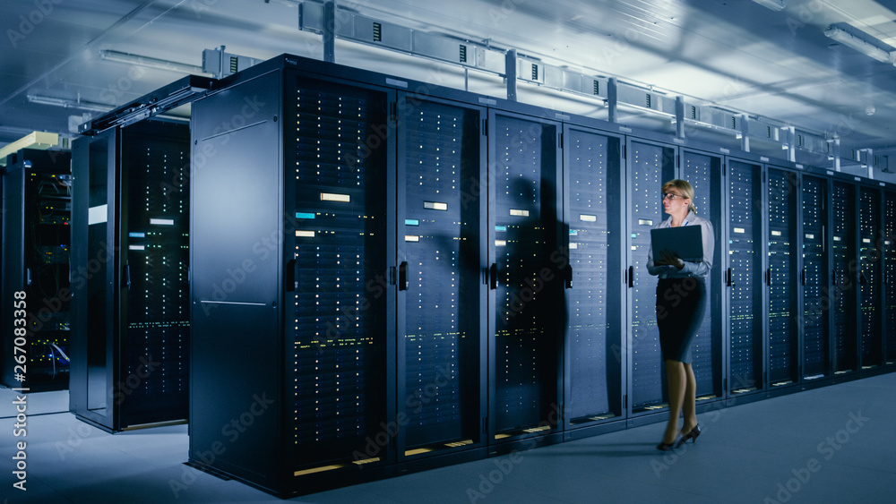 In Data Center: Female IT Technician Walks Alongside Row of Server Racks, Uses Laptop Computer ...