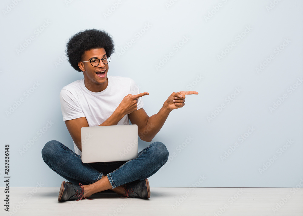 Young black man sitting on the floor with a laptop pointing to the side ...