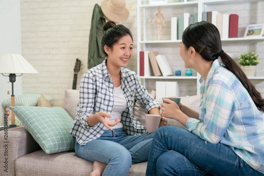 Happy asian japanese female friends at home sitting on couch. two young women with coffee ...