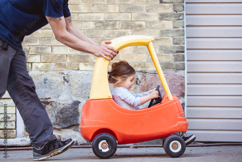 Portrait of a toddler girl being pushed by her daddy in a outdoor toy ...