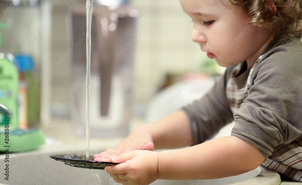 Little pretty child is washing dishes in kitchen. Nice toddler learns ...