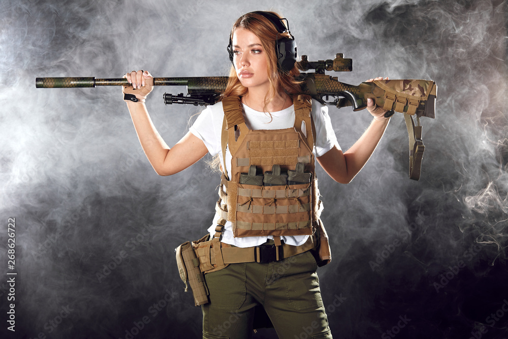 Skilled blonde female soldier with rifle in hands standing in military ...