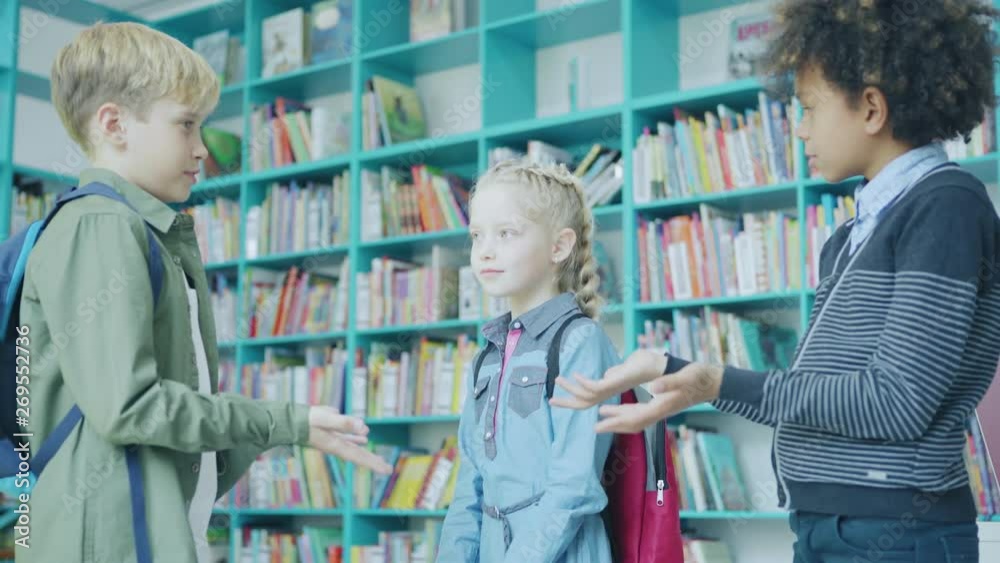 Three diverse students, two boys and girl, standing near book shelves ...
