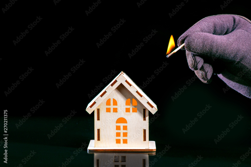 Hand with a burning match sets fire to the house model on black ...