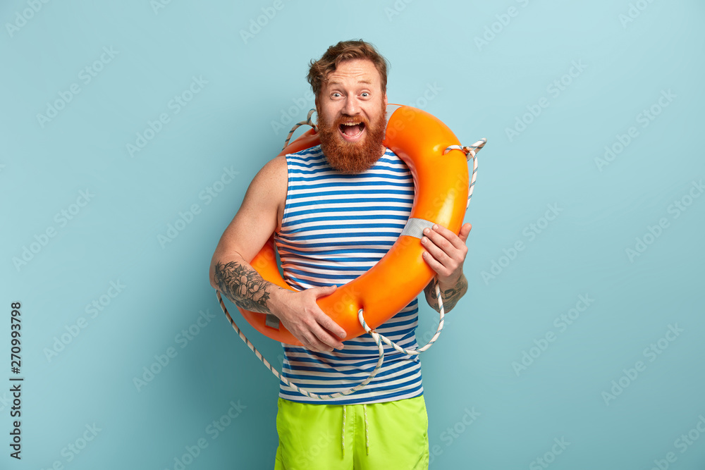 Overjoyed male lifeguard with tattoo, foxy beard, poses with inflated ...