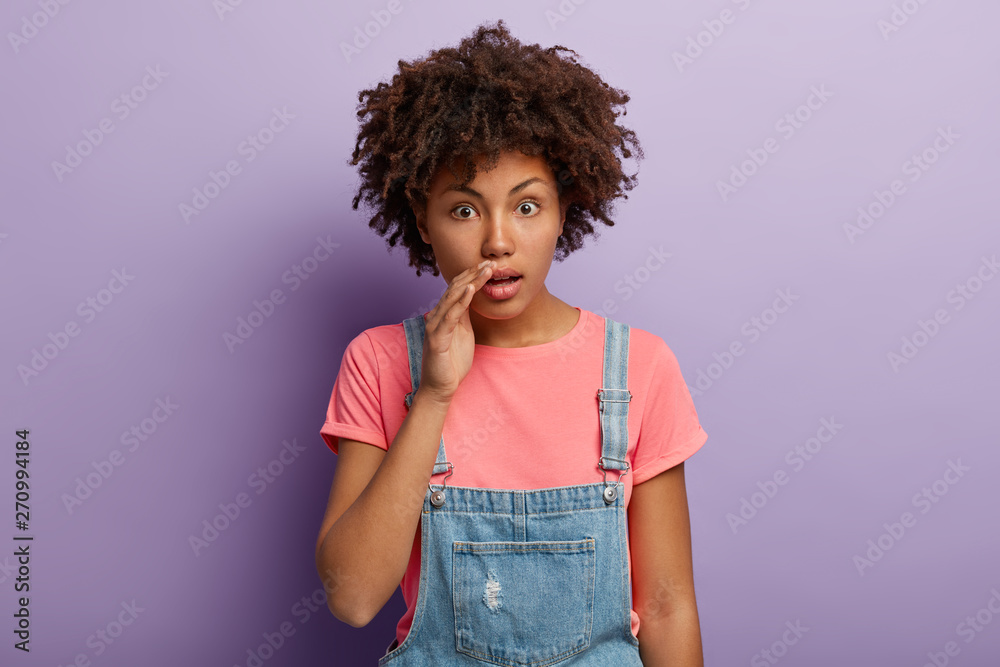 Talkative impressed dark skinned female keeps hand near mouth, tells ...